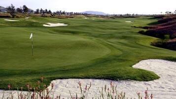 Um campo de golfe com bunkers de areia, uma haste de bandeira e uma cordilheira ao longe.