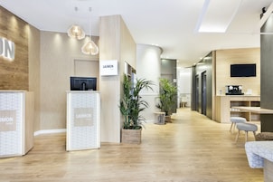Um saguão de hotel moderno com piso de madeira, balcão de recepção, plantas em vasos e uma área de cozinha ao fundo.