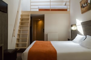 Quarto de hotel com beliche, escada de madeira, colcha branca e mesa de cabeceira com abajur.