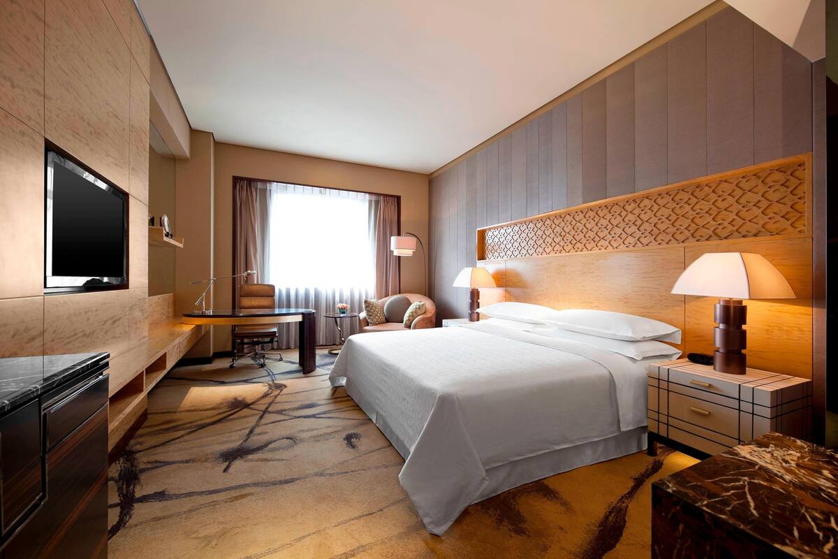 Quarto de hotel com cama, mesa de cabeceira, luminária, televisão e escrivaninha com cadeira.