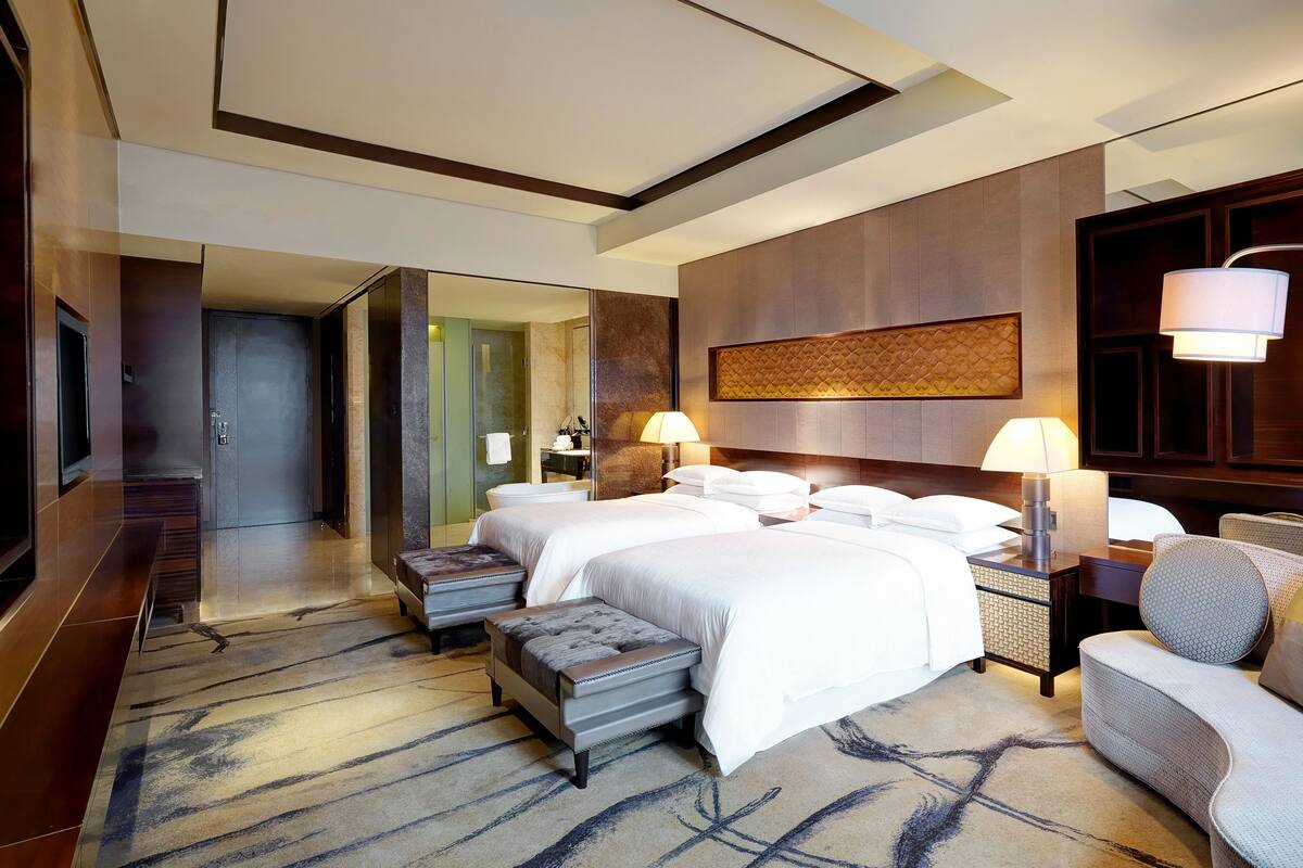 Quarto de hotel com duas camas, um sofá, uma televisão e um banheiro visível por uma porta aberta.