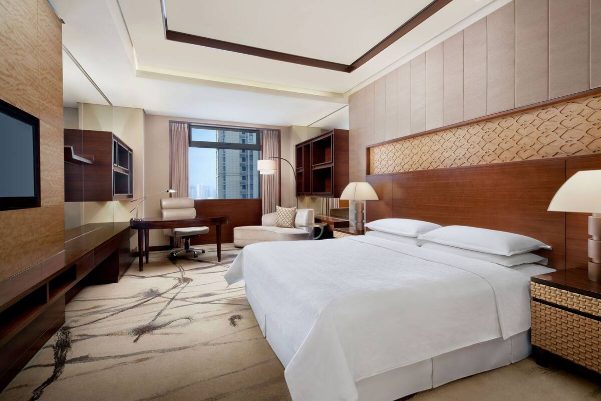 Quarto de hotel com uma cama grande, uma escrivaninha, uma cadeira, uma televisão e uma janela com vista para a cidade.