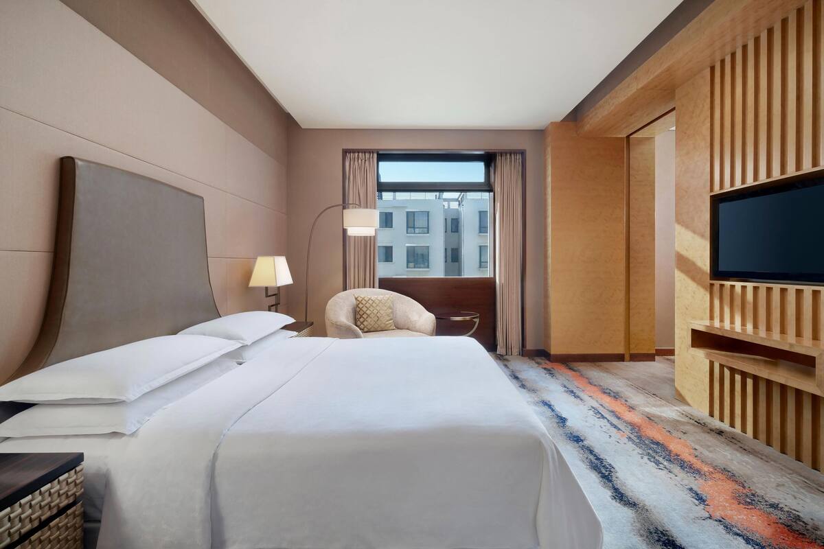 Quarto de hotel com uma cama grande, uma cadeira, uma televisão e uma janela com vista para edifícios.