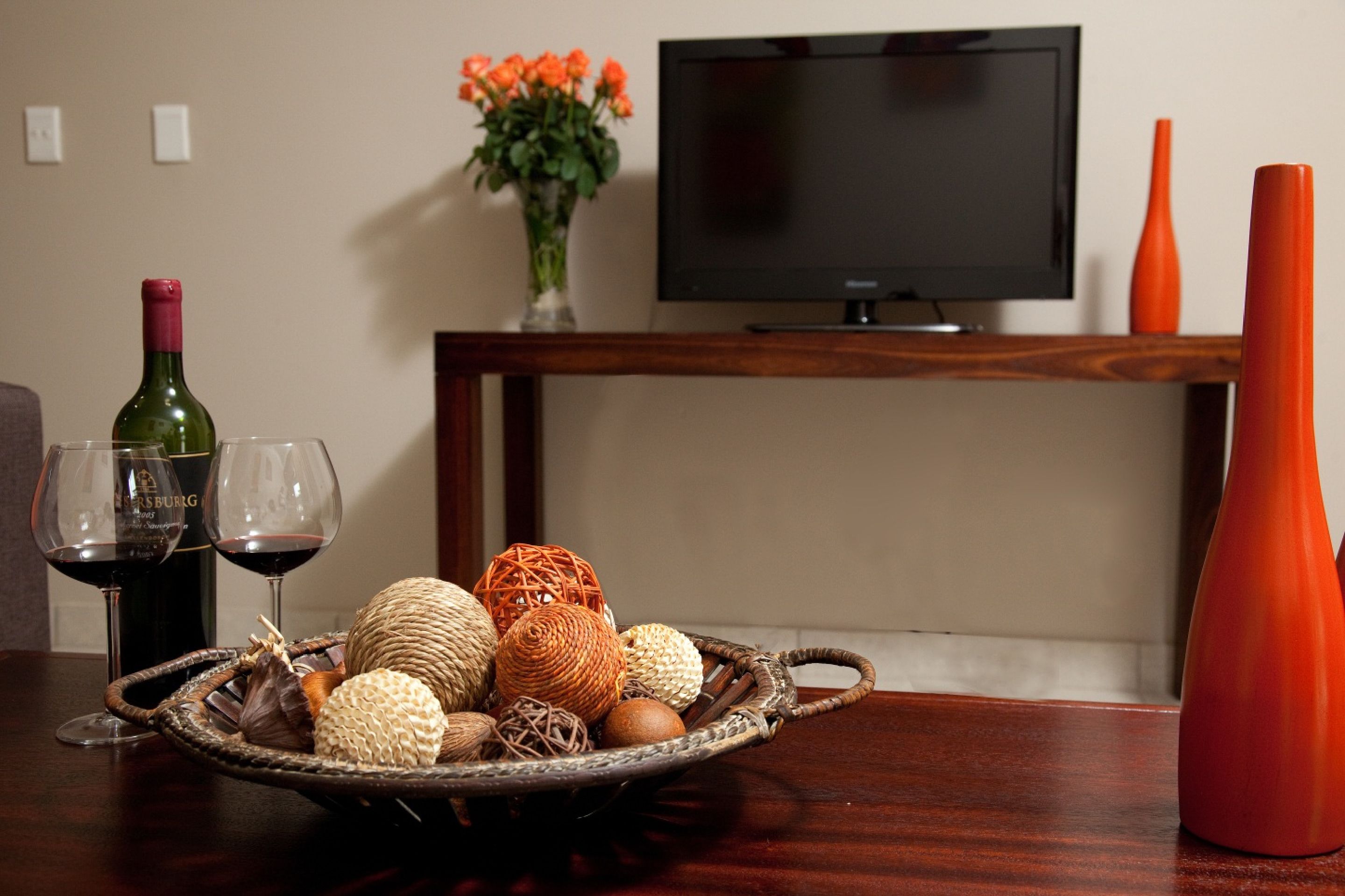 Mesa de jantar com cesta de alimentos, duas taças de vinho, uma garrafa de vinho e uma televisão ao fundo.