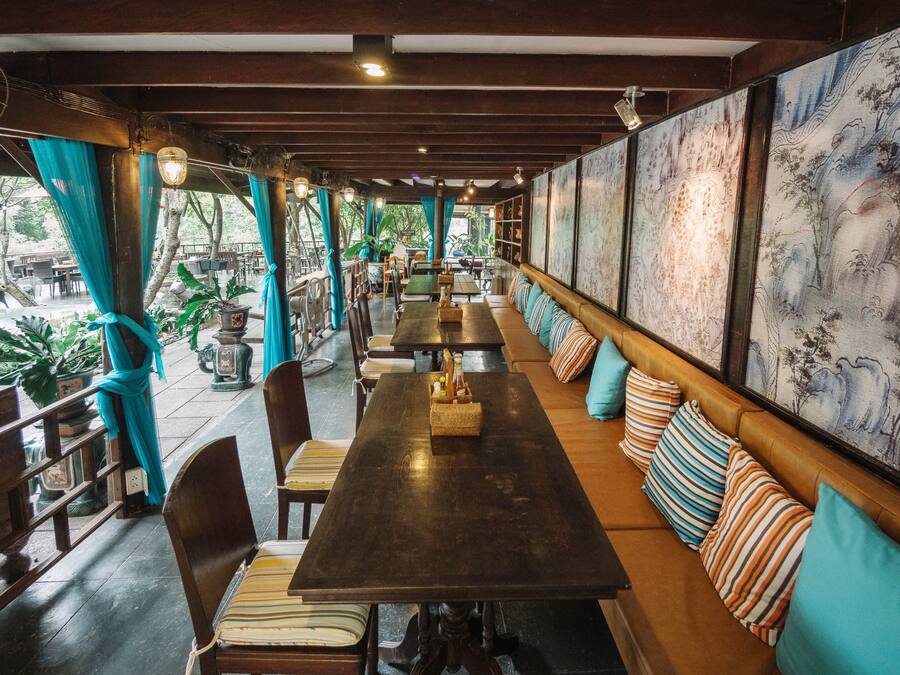 Um restaurante interno com mesas e bancos de madeira, plantas em vasos e obras de arte nas paredes.