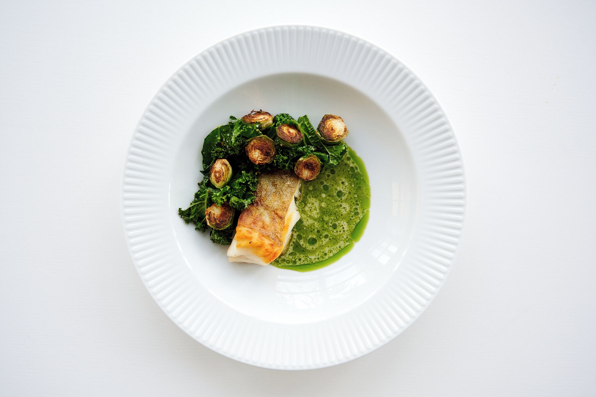 Um prato branco com um pedaço de peixe, brócolis de Bruxelas assados e um molho verde.
