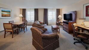 Quarto de hotel com sala de estar, mesa de jantar com cadeiras, sofá, televisão e escrivaninha com luminária.