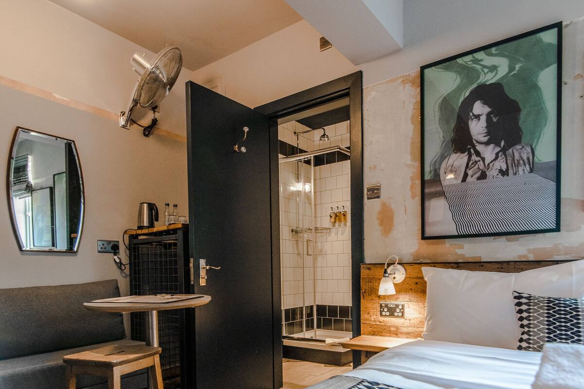 Quarto de hotel moderno com cama, área de estar com mesa e cadeira, espelho, ventoinha, chuveiro e um quadro na parede.