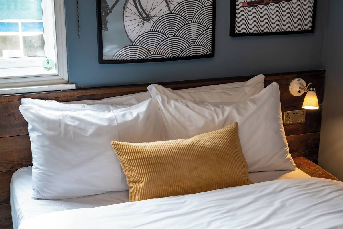 Cama bem arrumada com almofadas brancas e bege, cabeceira de madeira e luminária fixada na parede.