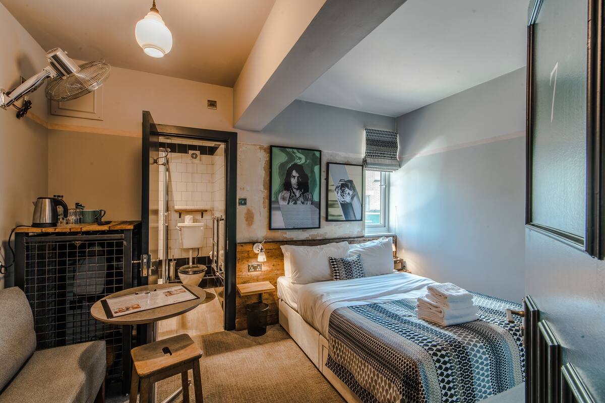 Quarto de hotel com uma cama, uma mesinha, uma cadeira, uma pia e uma janela.