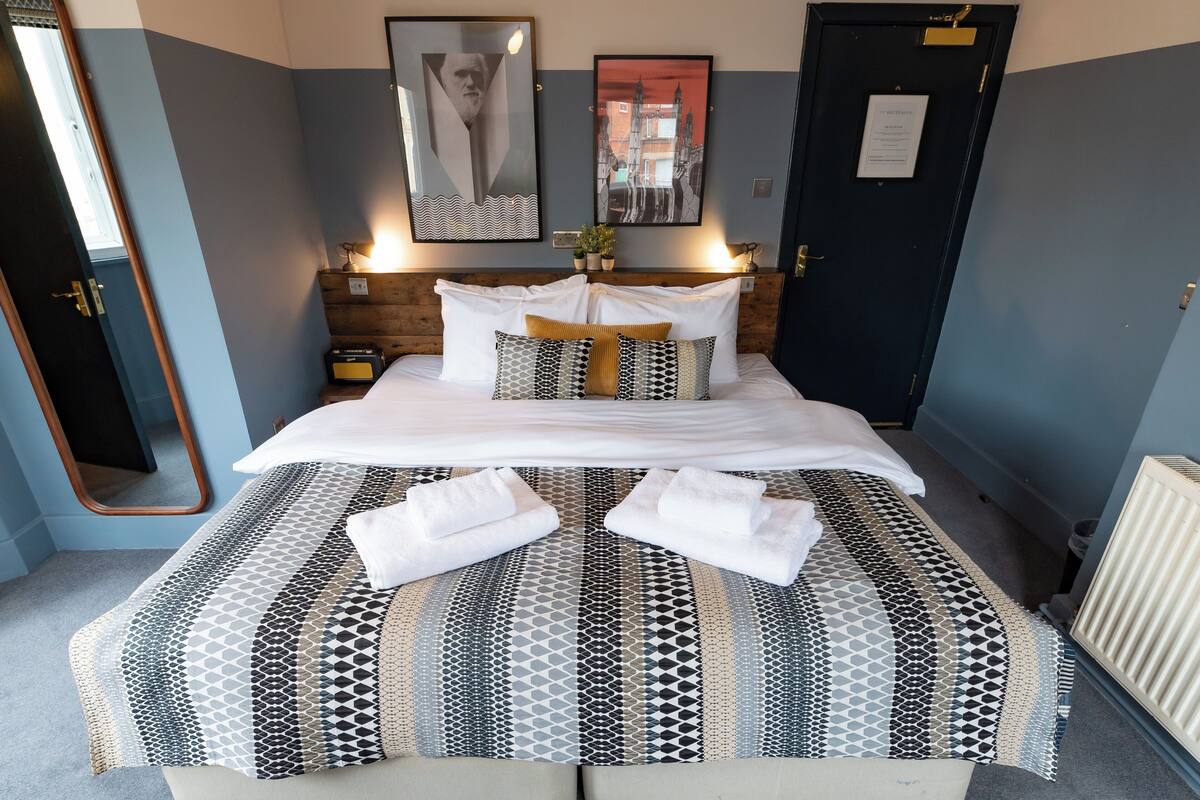 Cama bem arrumada com roupa de cama estampada, mesa de cabeceira com abajur e espelho à esquerda.