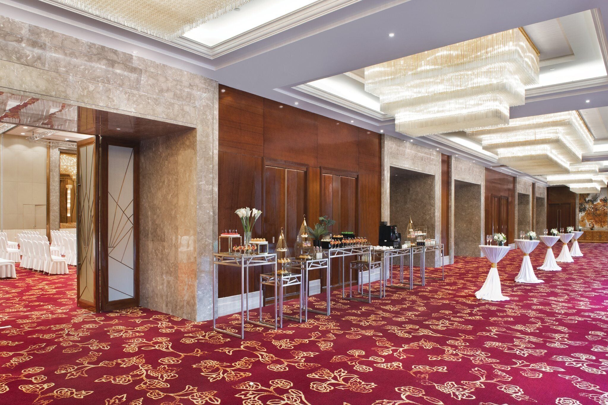 Uma sala de banquetes com fileiras de cadeiras brancas, mesas com centros de mesa florais e um tapete vermelho com padrão dourado.