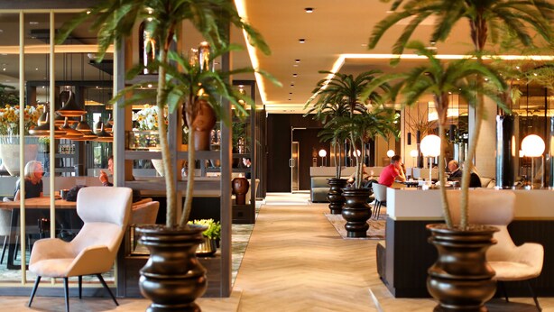 Um hall de hotel moderno, com um balcão de recepção central, áreas de estar e plantas em vasos.