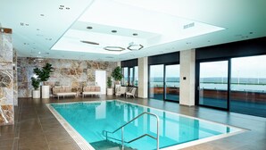 Piscina interior moderna com vista ampla para o mar através de grandes janelas.