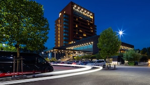 Um hotel moderno com entrada imponente, vários andares e área de estacionamento com veículos.