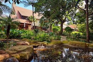 Um jardim tropical com lago, vegetação exuberante e um edifício com telhado vermelho.