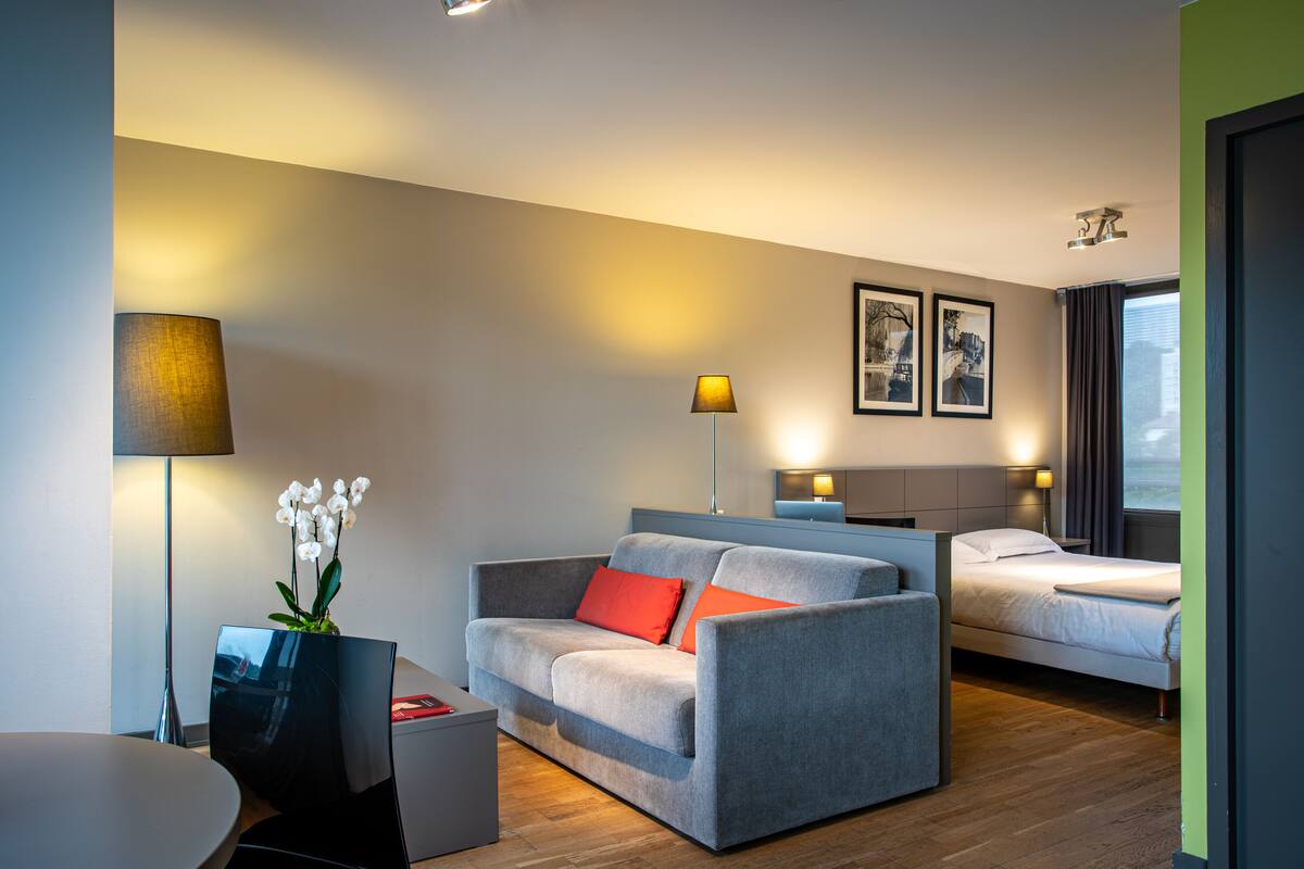 Quarto de hotel moderno com um sofá cinzento, uma cama, uma escrivaninha e uma luminária.