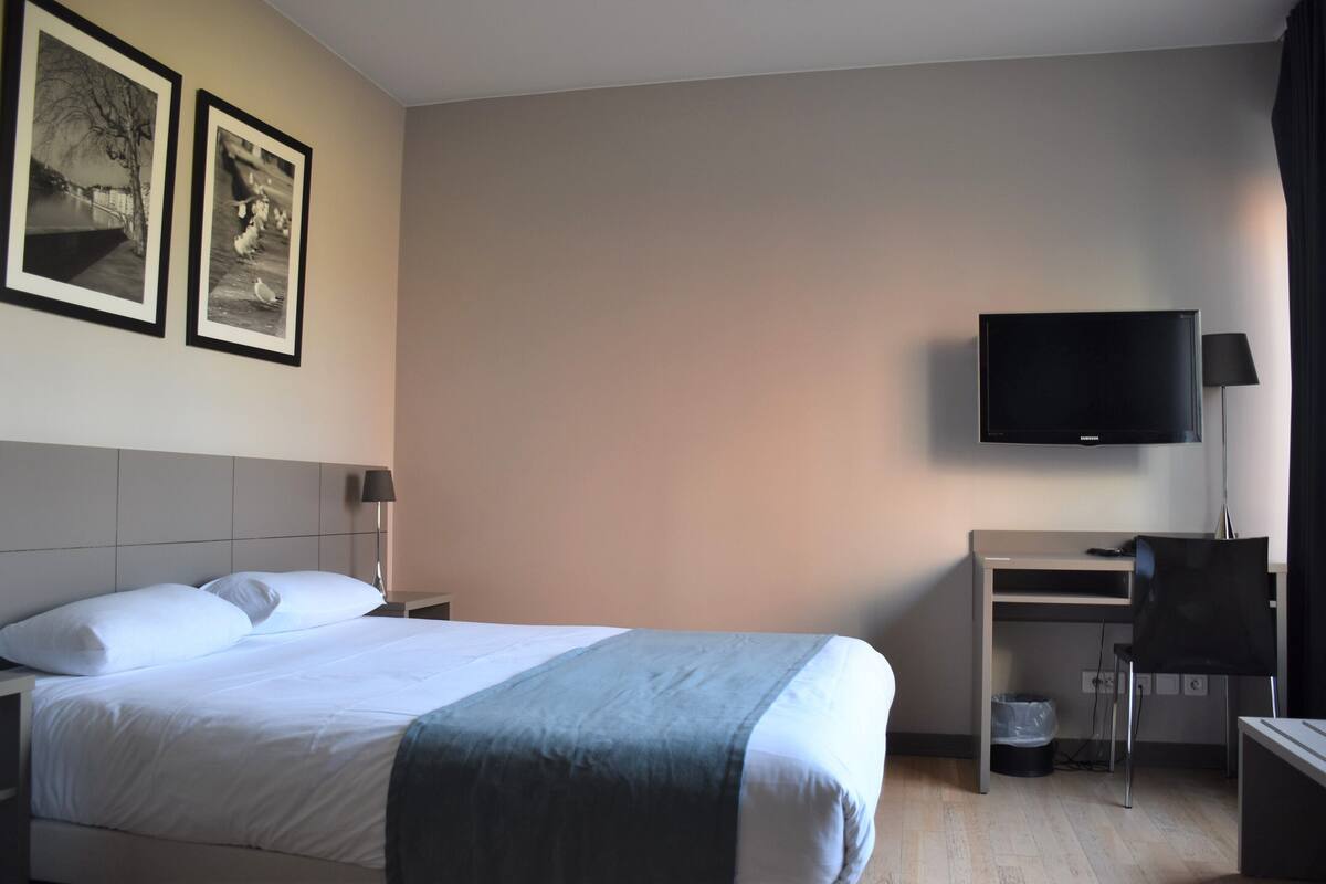 Quarto de hotel com uma cama, uma mesa, uma televisão e duas imagens emolduradas na parede.