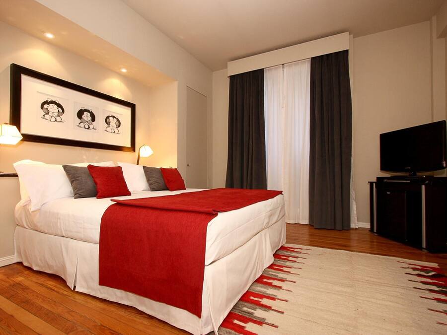 Uma cama bem arrumada com um cobertor vermelho, um quadro emoldurado acima e uma televisão sobre um suporte à direita.
