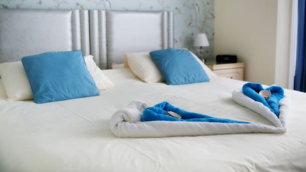 Quarto de hotel com uma cama, duas almofadas, mesa de cabeceira e uma luminária.