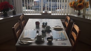 Mesa de jantar posta para quatro pessoas, cadeiras, janela com persianas e vista para carros estacionados e edifícios lá fora.