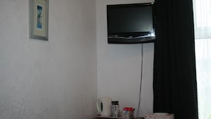 Quarto com televisão de tela plana na parede, chaleira branca sobre um tabuleiro e pequena mesa com uma caixa e uma xícara.