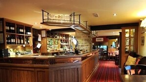 Uma área de bar com balcão de madeira, prateleiras com garrafas e tapete xadrez vermelho.