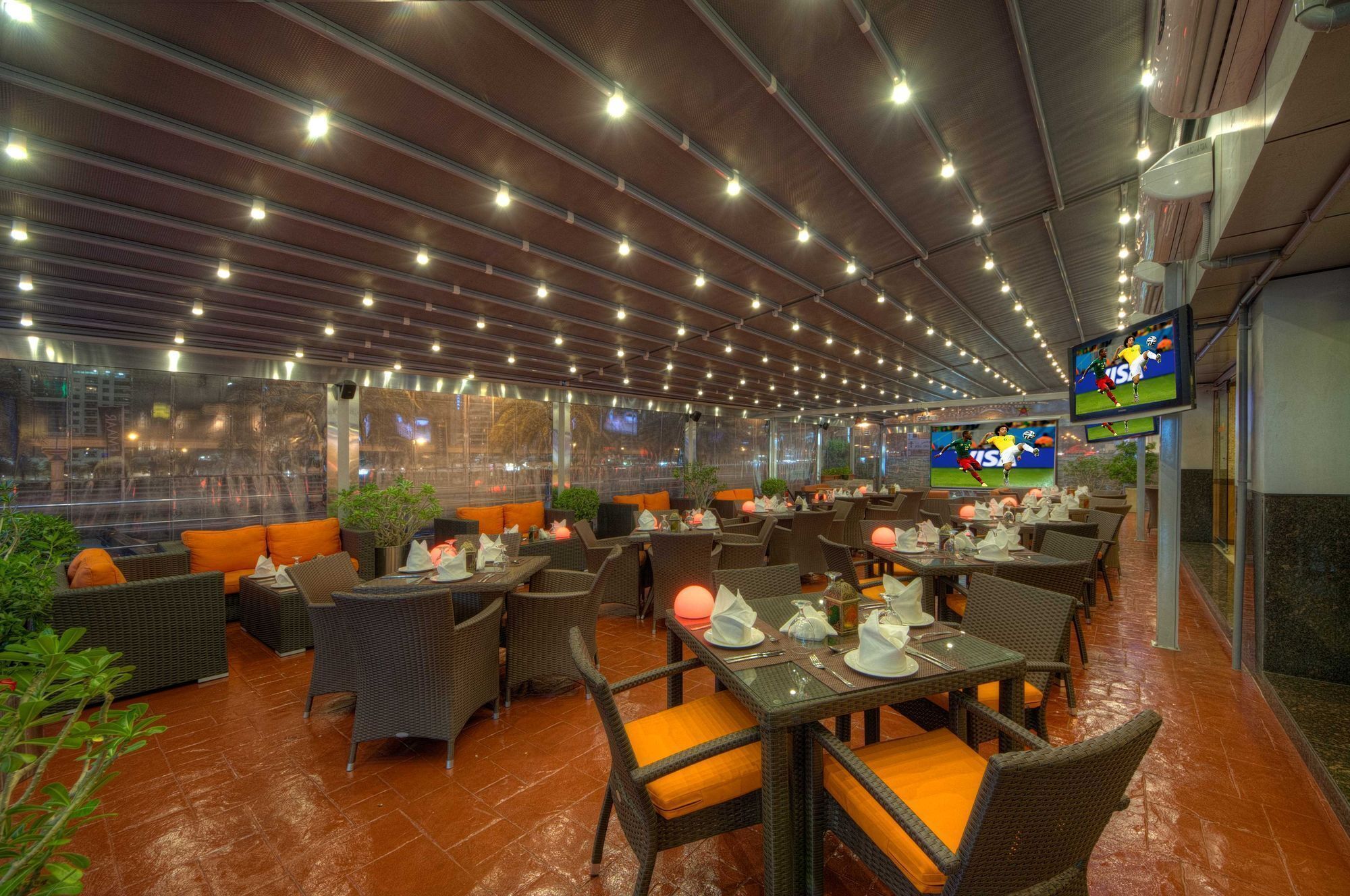 Restaurante interno com mobiliário de vime, mesas montadas com louças brancas e uma grande televisão exibindo um evento esportivo.