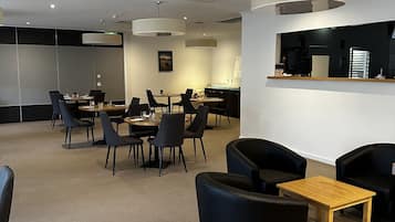 Um hall de hotel moderno com área de refeições, sofás de couro preto, uma mesa de madeira e um balcão de recepção.