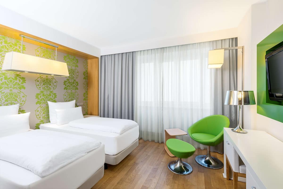 Quarto de hotel com duas camas, uma escrivaninha, uma cadeira, uma televisão e uma janela com cortinas.