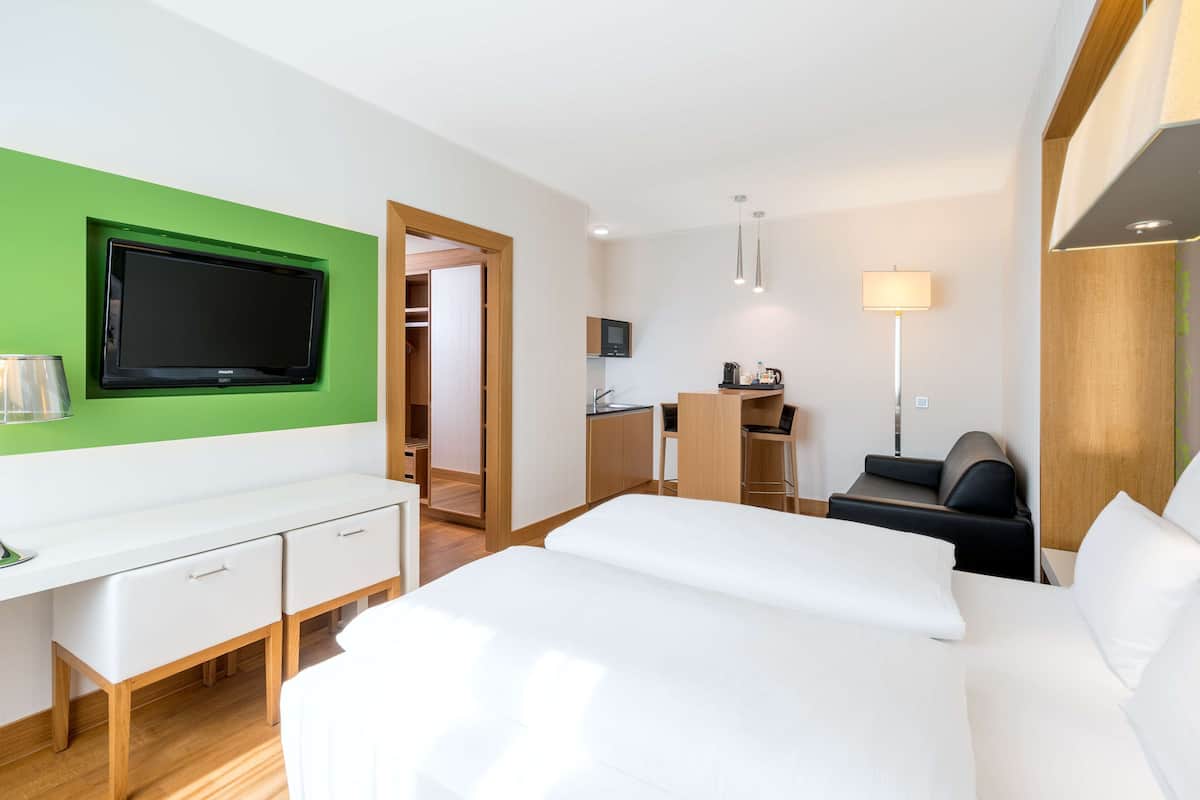 Quarto de hotel moderno com uma cama, uma mesa de escritório, uma TV e uma cozinha compacta.