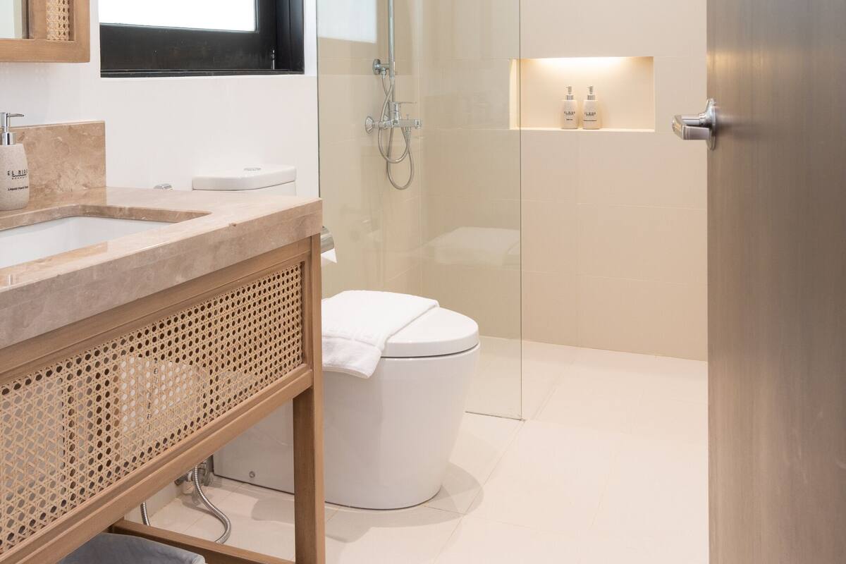 Uma casa de banho moderna com chuveiro separado por vidro, sanita, lavatório com espelho e prateleira de madeira.