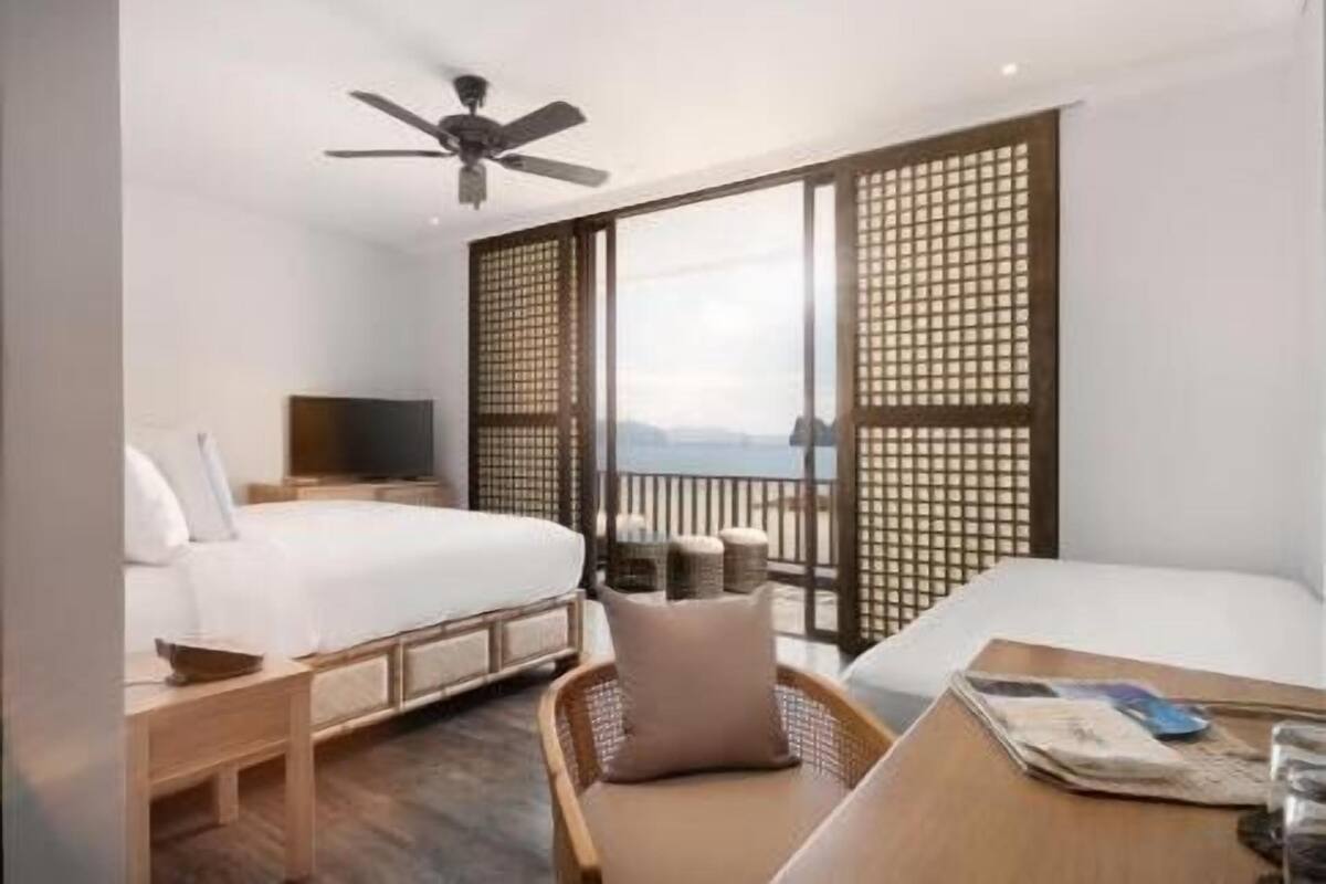 Quarto de hotel com cama, escrivaninha, cadeira, ventilador de teto e vista para o mar.
