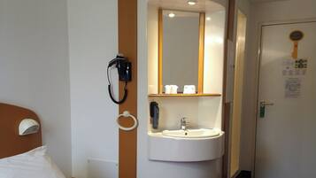 Quarto de hotel com cama, colcha vermelha, parede branca, banheiro com pia e espelho, e porta com fechadura eletrônica.