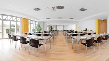 Sala de conferências com várias mesas e cadeiras, janelas amplas e um projetor.