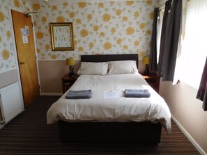 Quarto de hotel com cama, duas mesas de cabeceira, um criado-mudo, luminária, quadro na parede e uma porta.