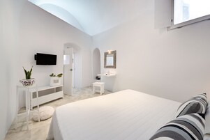 Quarto moderno com uma cama grande, TV fixada na parede, um pequeno banco branco e um vaso com planta.