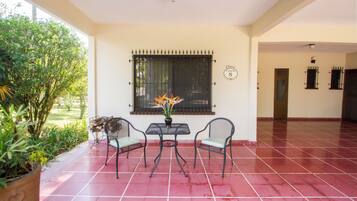 Uma área de varanda coberta com piso de azulejos, uma mesa com cadeiras e um vaso de planta.