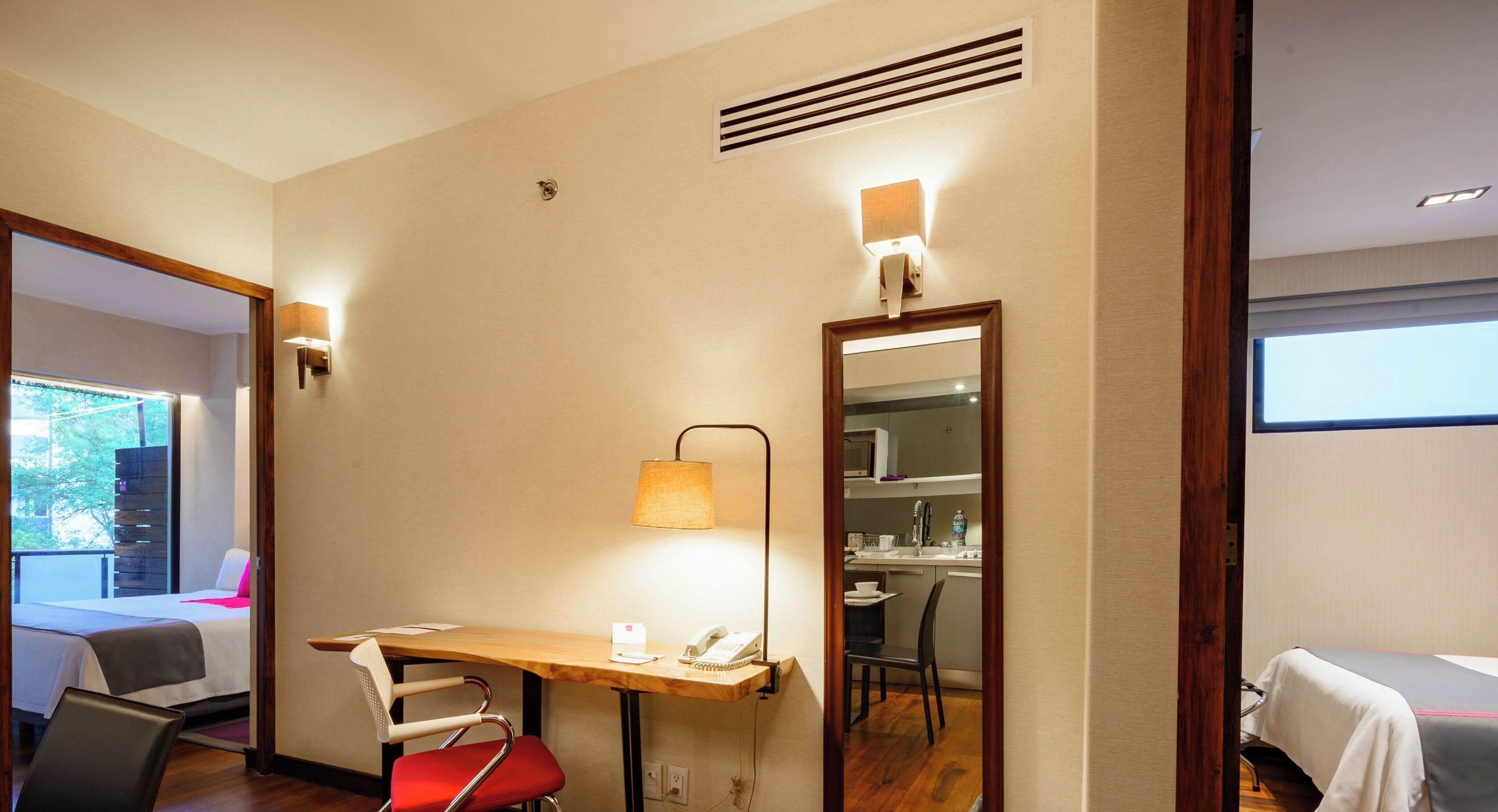 Quarto de hotel com escrivaninha de madeira, cadeira vermelha, mesa de cabeceira, espelho e janela.