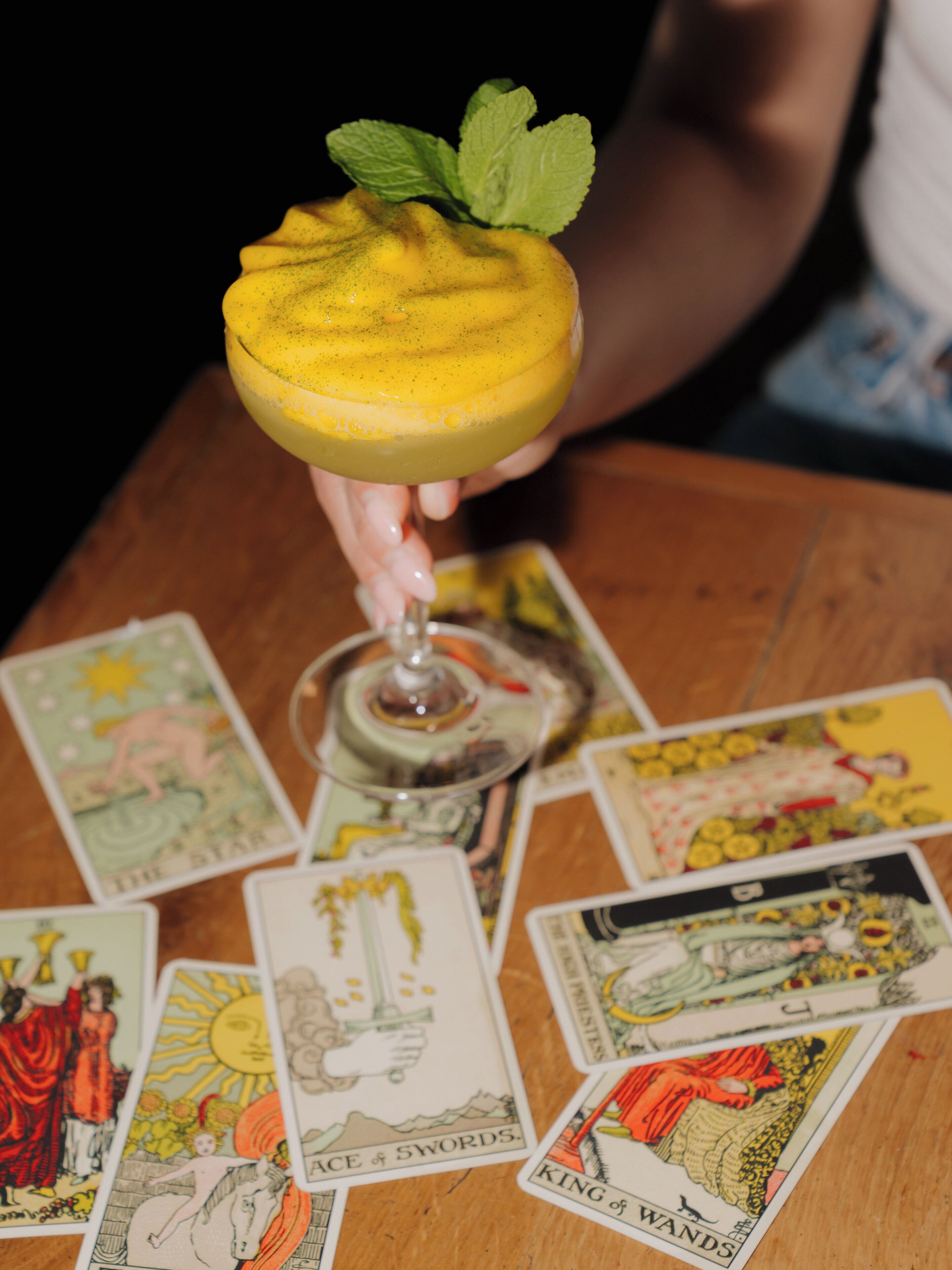 Uma mão segura um coquetel com um ramo de hortelã, cercado por cartas de tarot sobre uma mesa de madeira.