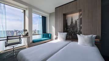 Quarto de hotel com uma cama grande, uma mesa com cadeira, um assento junto à janela e vista para a cidade.