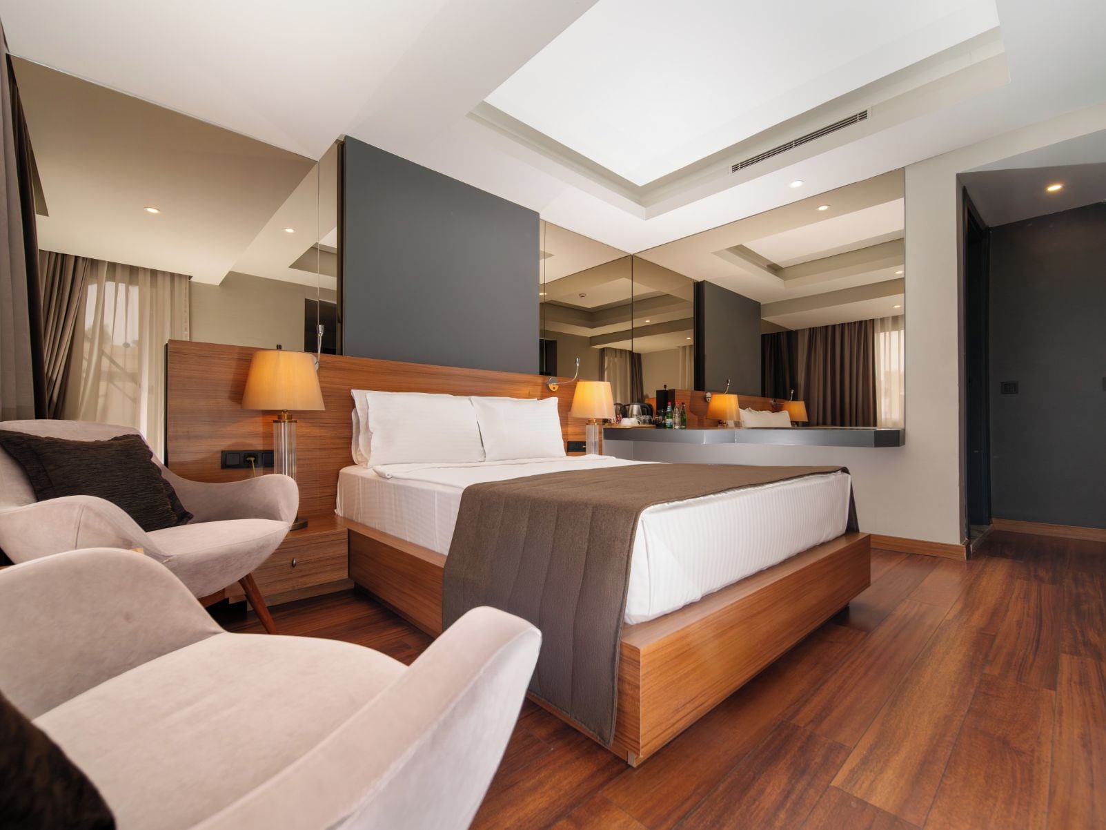 Quarto de hotel moderno com uma cama, dois poltronas, uma escrivaninha e um espelho grande.
