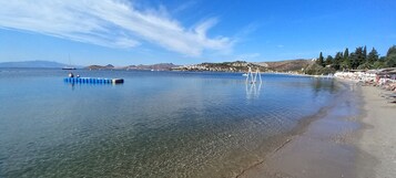 Uma praia calma com águas cristalinas, um cais e uma linha de costa distante com edifícios e colinas.
