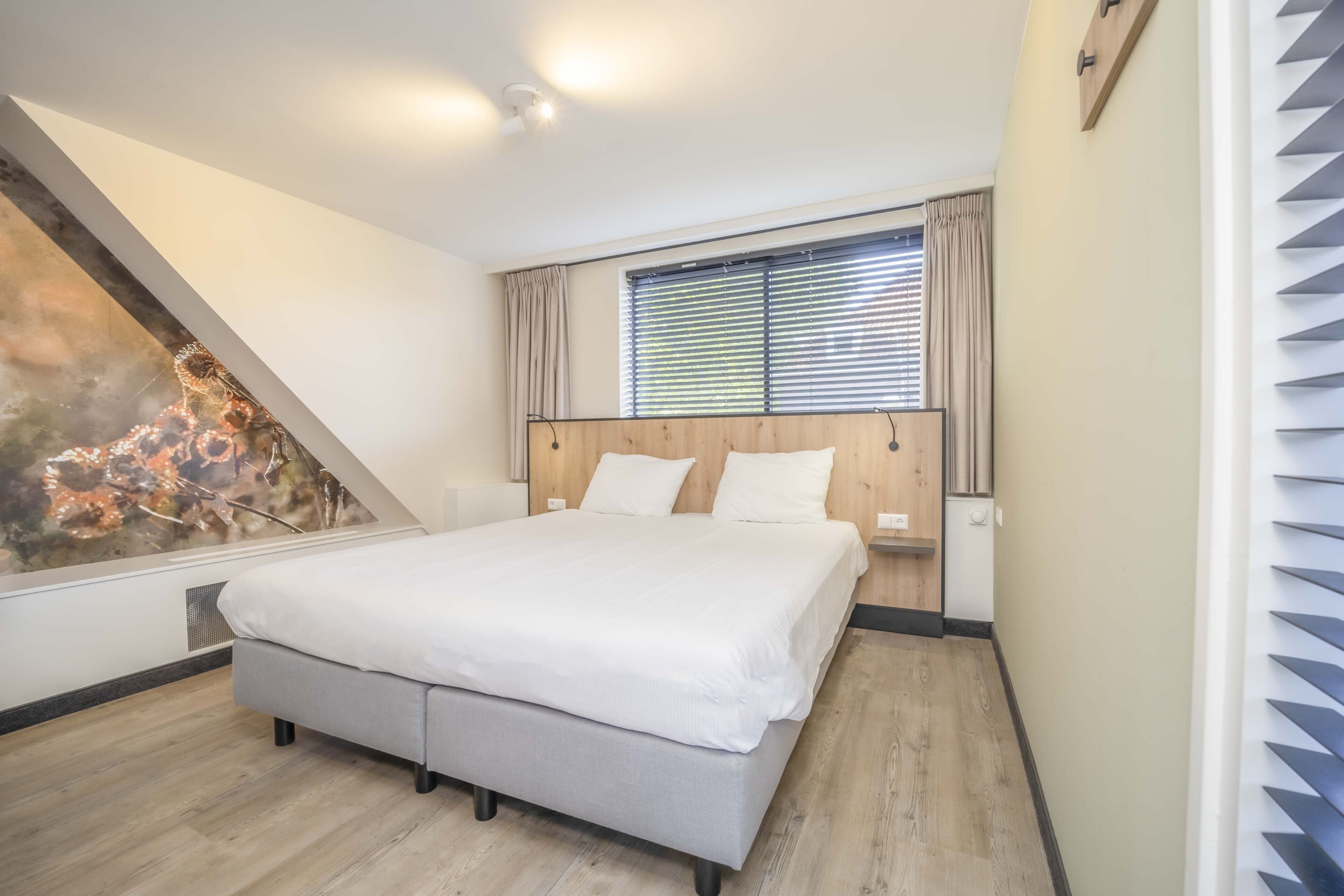 Quarto com cama grande, cabeceira de madeira, janela com persianas e vista parcial de um mural na parede.