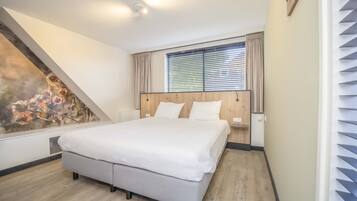 Quarto com cama grande, cabeceira de madeira, janela com persianas e vista parcial de um mural na parede.