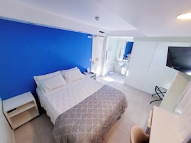 Quarto de hotel com uma cama, escrivaninha, cadeira, televisão e banheiro visível por uma porta aberta.