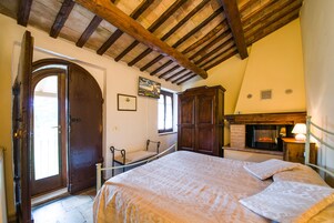 Quarto com cama grande, vigas de madeira no teto, lareira e porta que dá para o exterior.