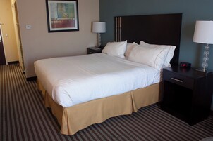 Quarto de hotel com uma cama grande, uma mesa de cabeceira com abajur, um relógio digital e um quadro na parede.