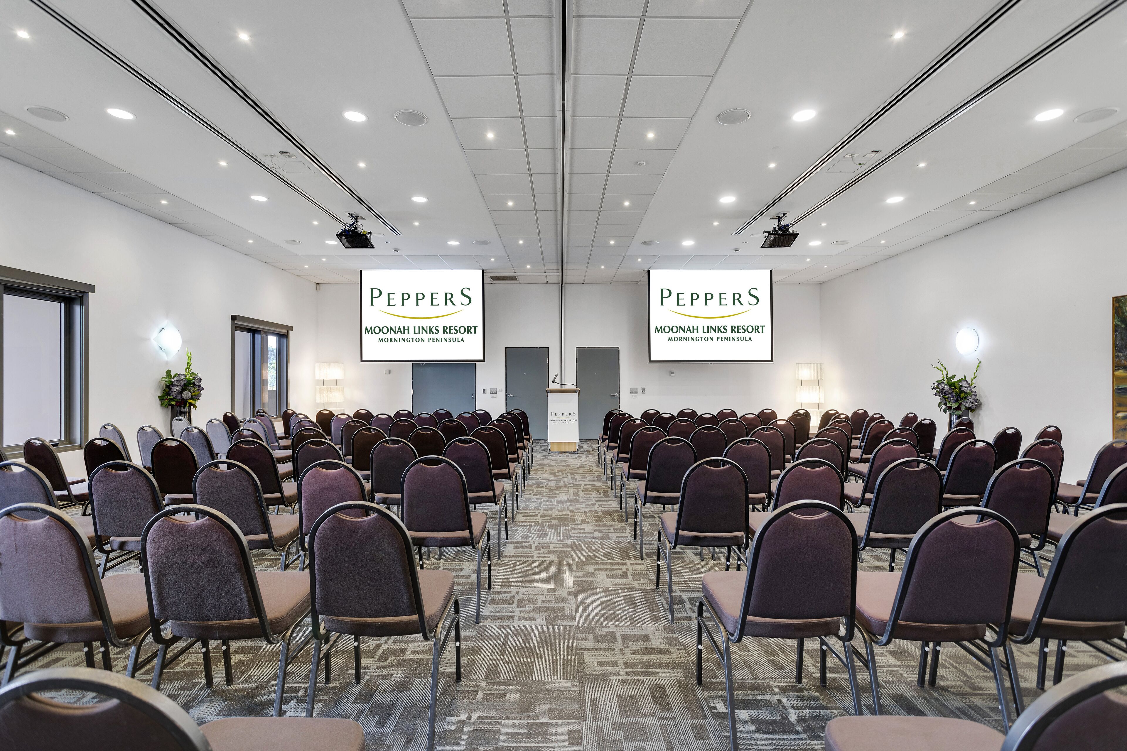 Uma sala de conferências com fileiras de cadeiras, piso coberto por carpete e iluminação no teto.