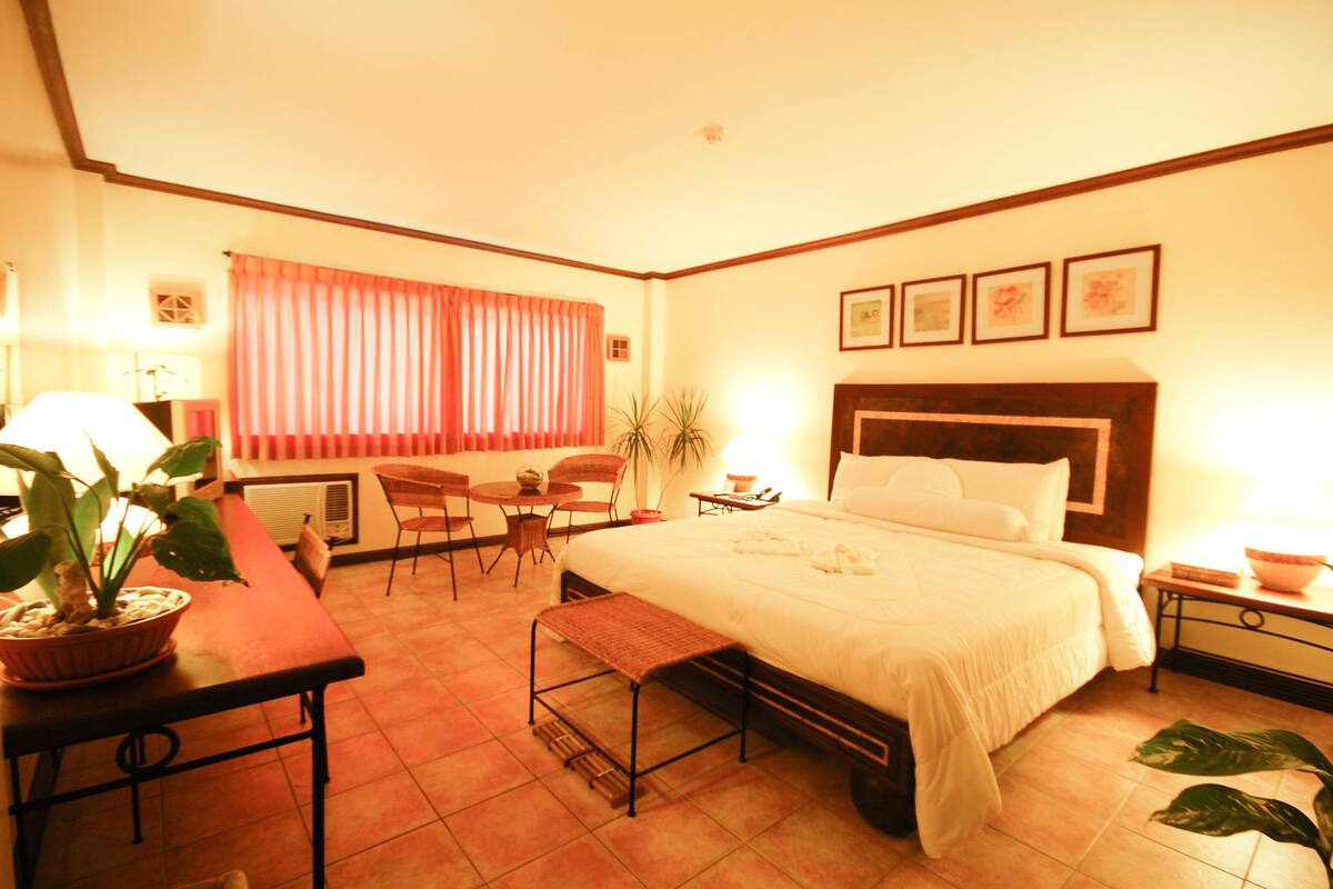 Quarto de hotel com cama, escrivaninha, cadeira, mesa, planta em vaso, janela com cortinas e quadros nas paredes.